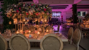 Salão de banquetes com longas mesas preparadas para um evento formal, decorado com arranjos florais e velas.