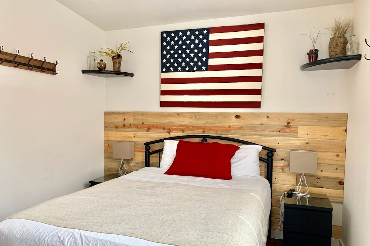 Quarto com cabeceira de madeira, cama com roupa de cama branca e vermelha, duas mesas de cabeceira com luminárias e uma bandeira americana na parede.