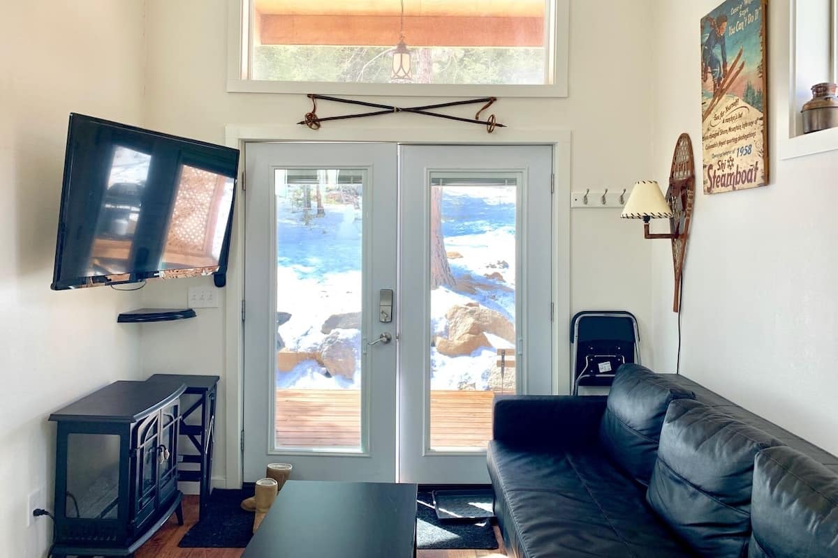 Uma sala de estar com televisão, um sofá de couro preto, uma mesa de madeira e uma porta que dá para uma área externa coberta de neve.