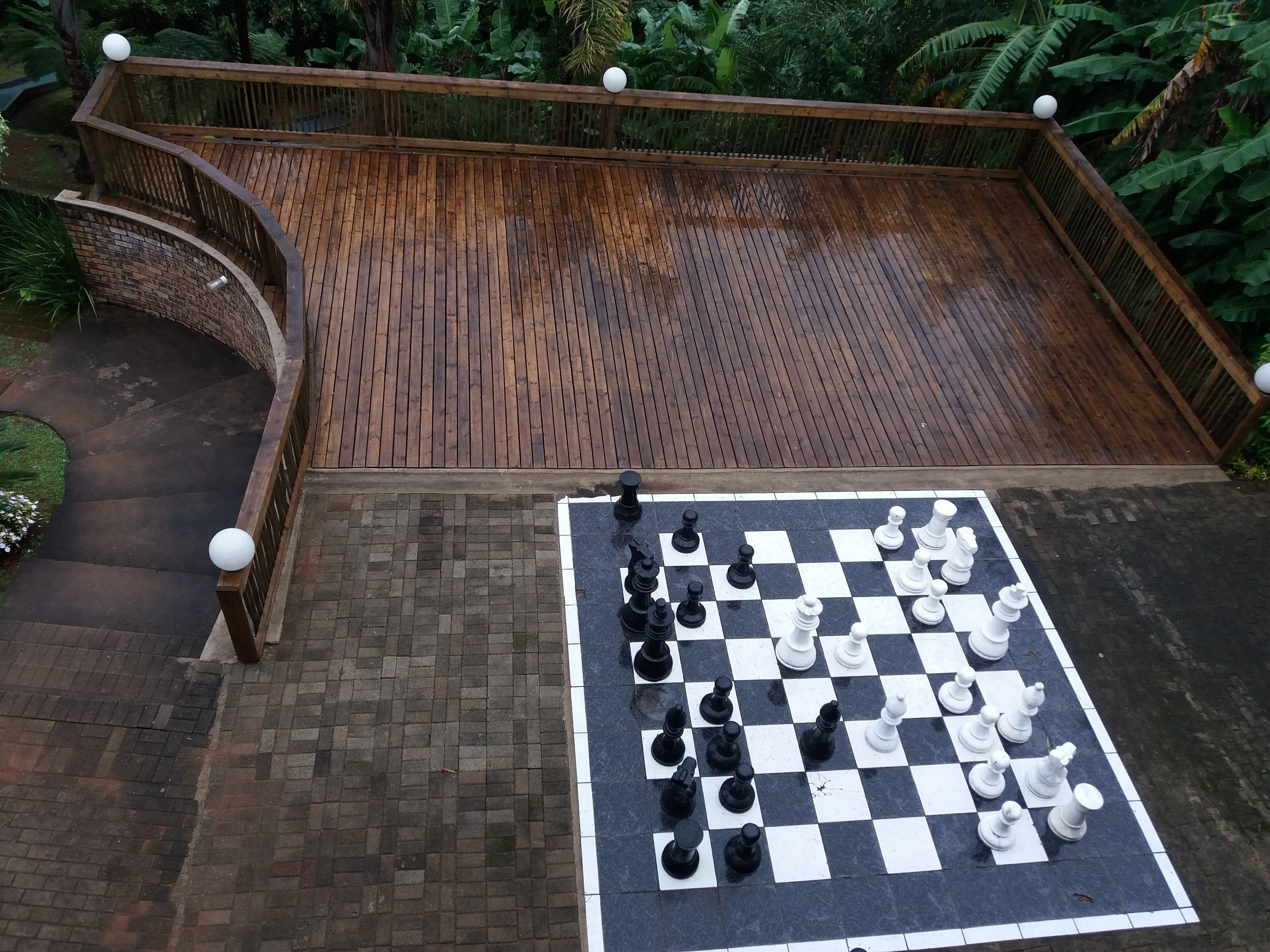 Um deck de madeira com um tabuleiro de xadrez disposto numa área pavimentada, rodeada por vegetação.