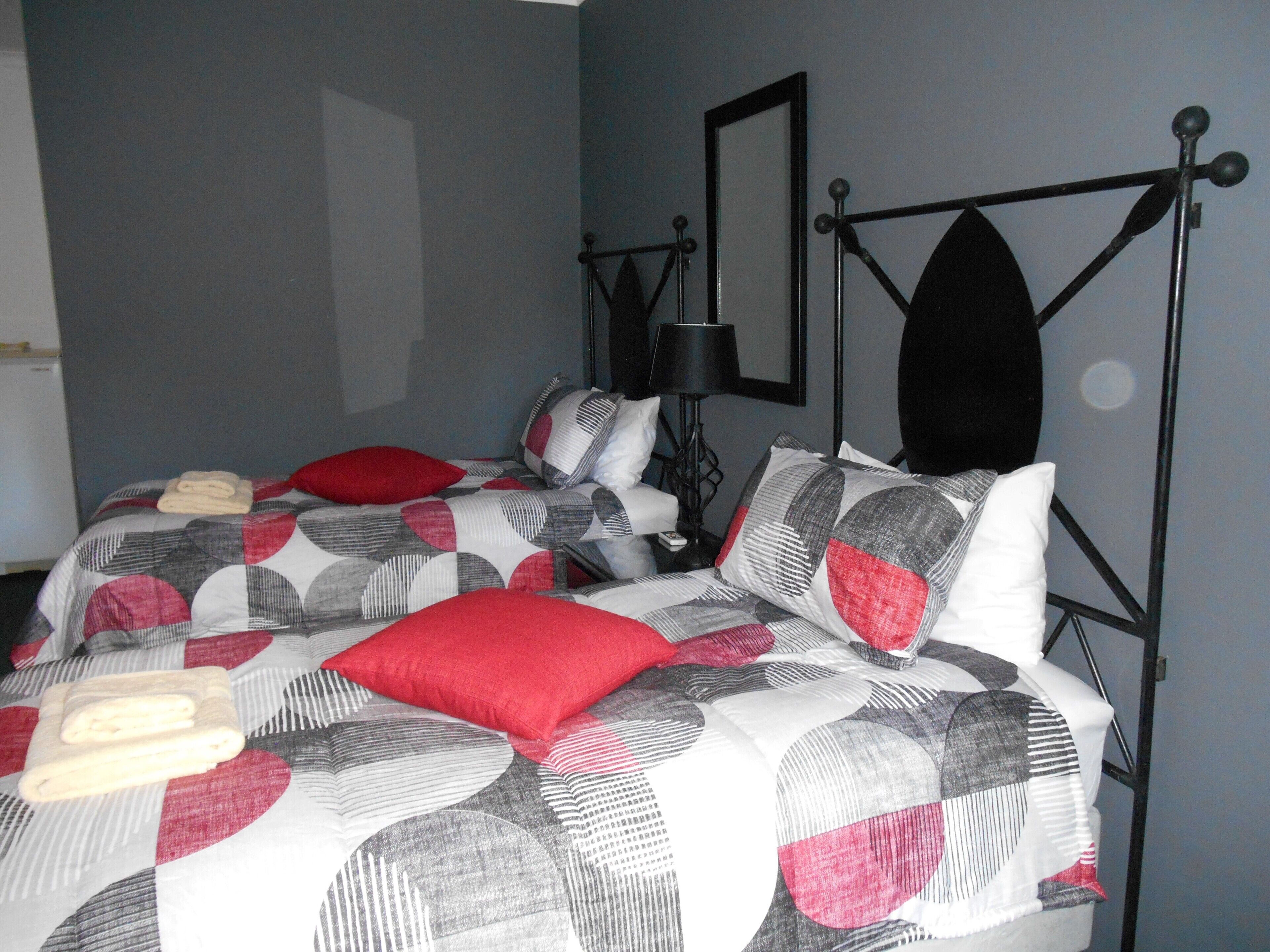 Cama bem arrumada com colcha estampada, um travesseiro vermelho e uma luminária preta ao lado.