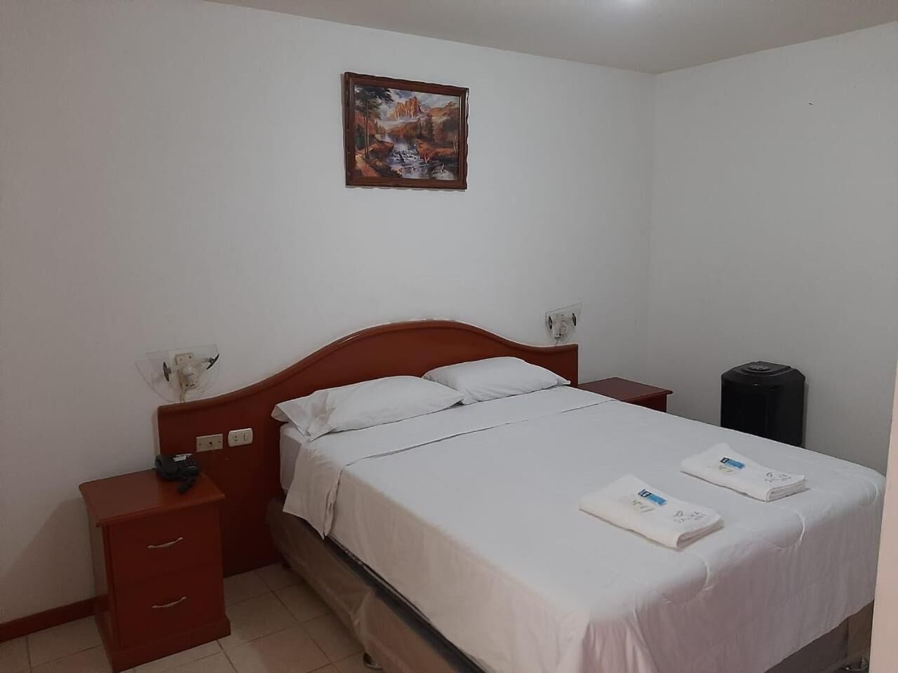 Quarto individual com uma cama de solteiro, cabeceira de madeira, roupa de cama branca, mesa de cabeceira, um criado-mudo, um quadro pendurado na parede e uma lixeira preta.