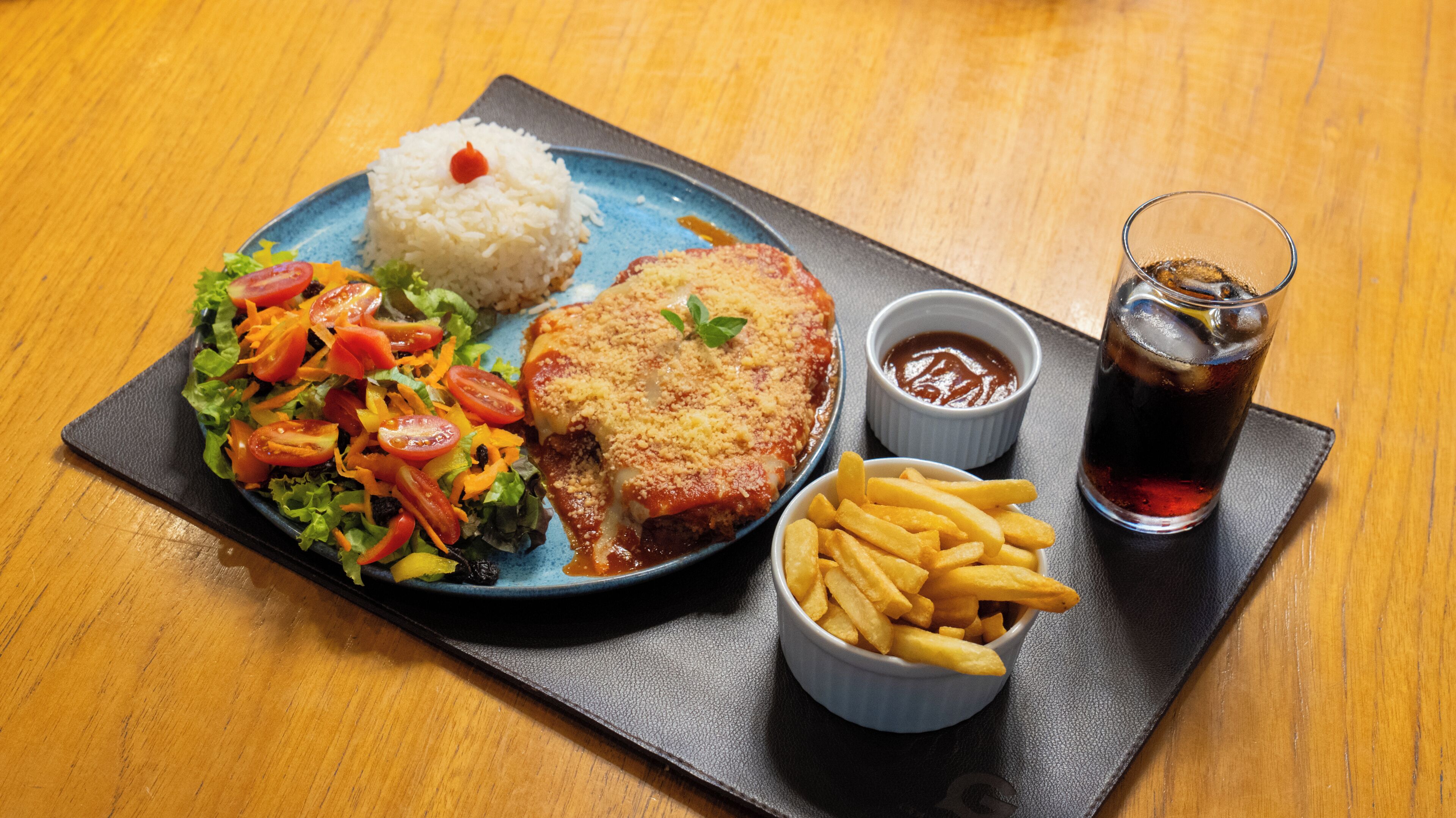 Um prato com salada, escalope empanado, arroz, batatas fritas e um copo de cola sobre uma mesa de madeira.