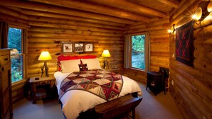 Um quarto de cabana de tronco com uma cama, duas luminárias de cabeceira, um colcha, um banco e uma pequena mesa.