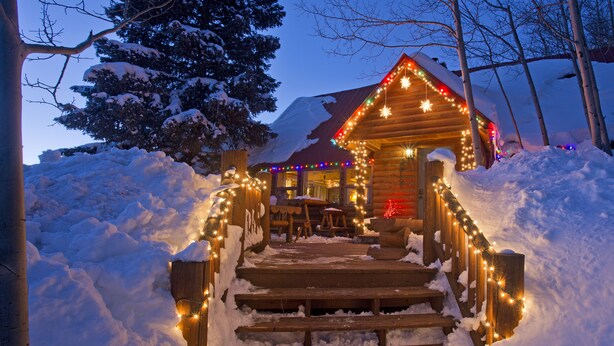 Uma cabana de madeira com varanda coberta de neve e degraus que levam à entrada, rodeada por neve e árvores.
