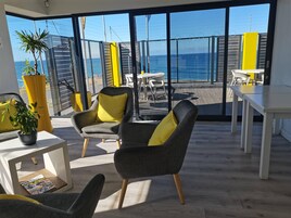 Um interior moderno com janelas amplas, vista para o mar e uma área de estar com cadeiras e mesa.