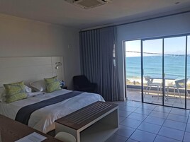 Quarto de hotel com cama, escrivaninha, cadeira e vista para o oceano através de amplas janelas.