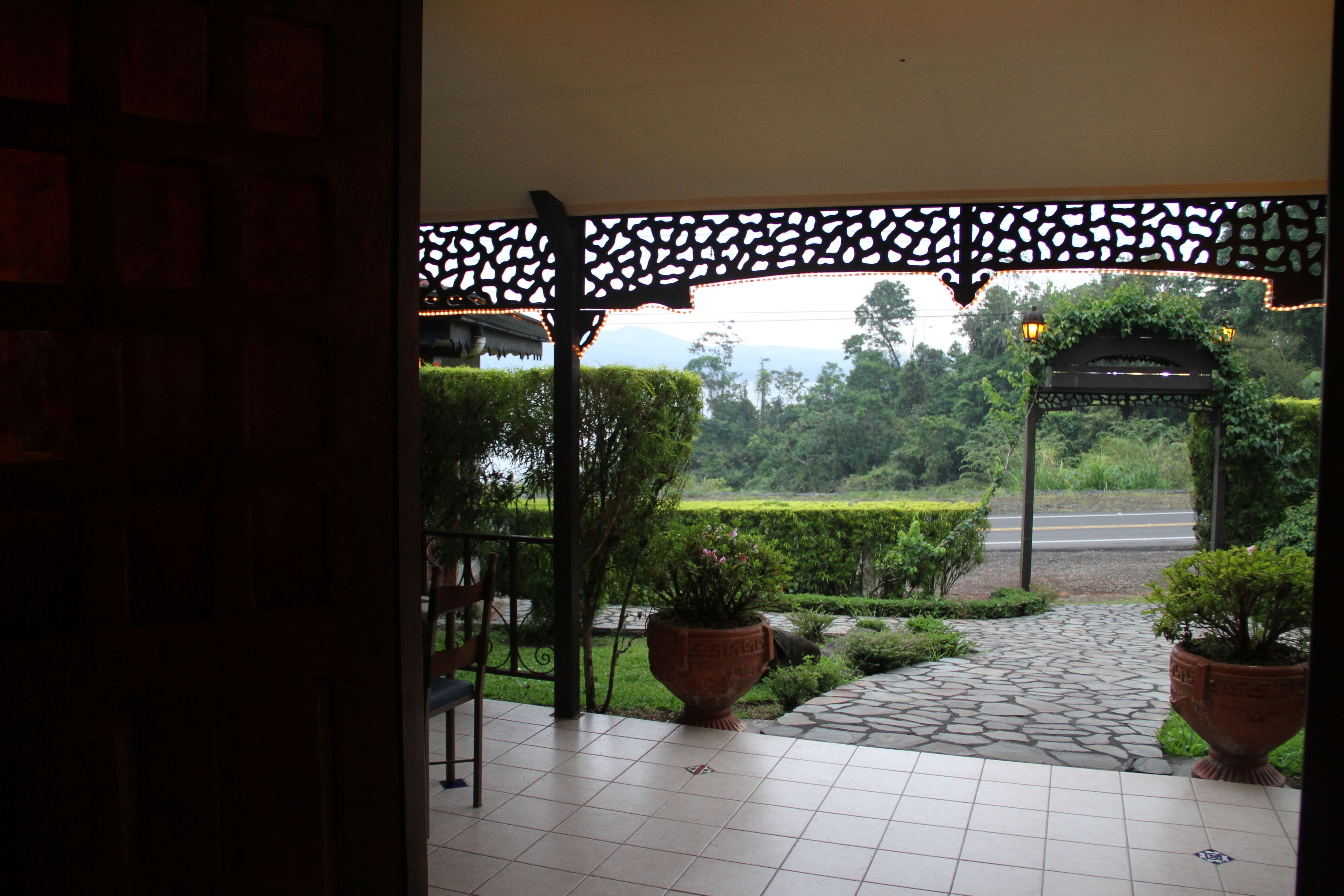 Visão através de uma porta de madeira para um jardim com caminho de pedra, plantas em vasos e um gazebo.
