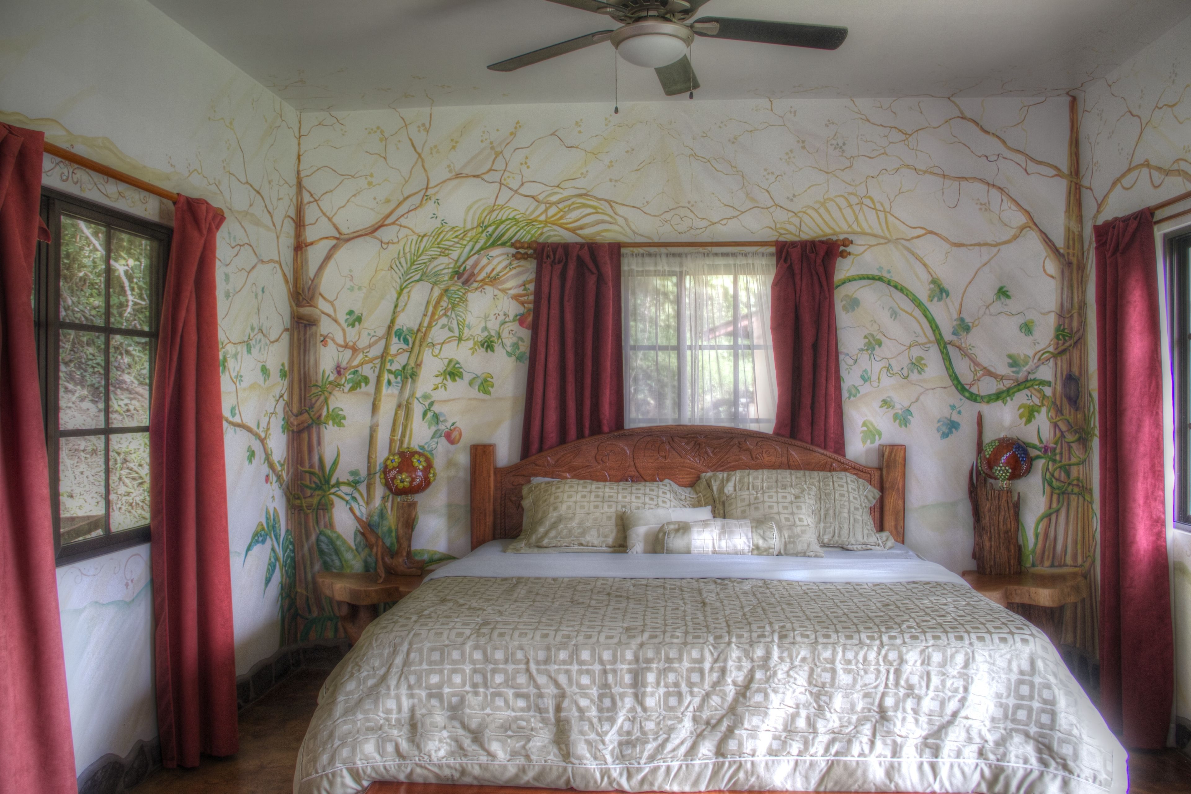 Quarto com uma cama grande, cortinas vermelhas, ventilador de teto e um mural de árvores e plantas.