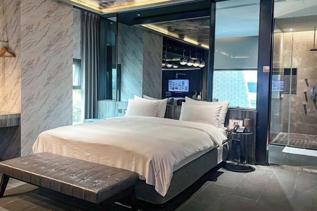 Quarto de hotel moderno com uma cama grande, um banco, televisão e área de chuveiro de vidro.