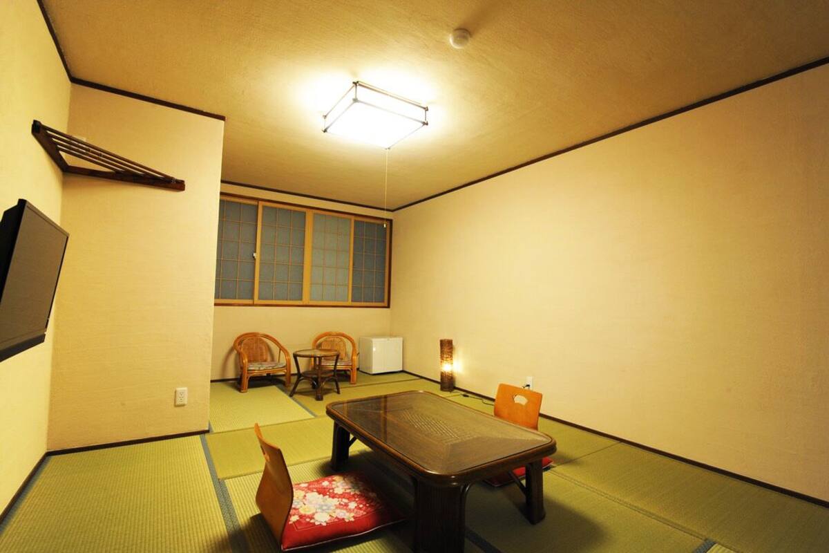 Um quarto japonês tradicional com tatames, uma mesa baixa, duas cadeiras, uma televisão e uma porta de correr.