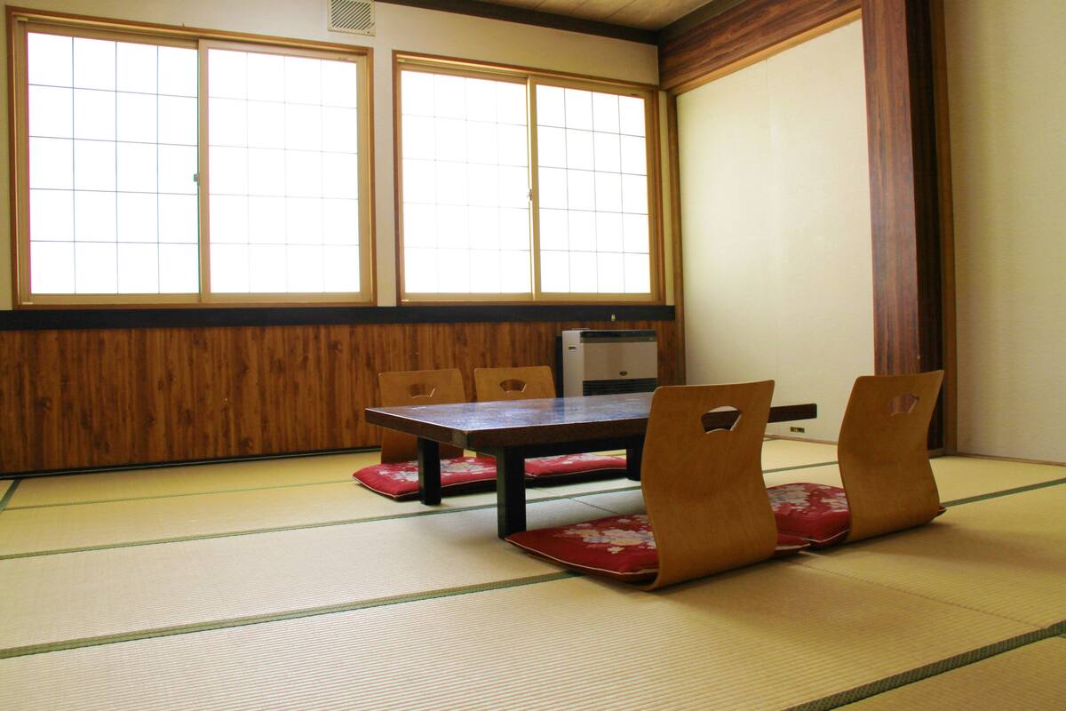 Um espaço com tatames, uma mesa baixa e quatro cadeiras, janelas amplas e um aparelho de ar condicionado tradicional japonês.