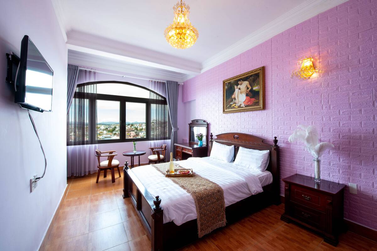 Quarto com uma cama grande, televisão fixada na parede, mesa de cabeceira de madeira, cômoda e janela com vista.