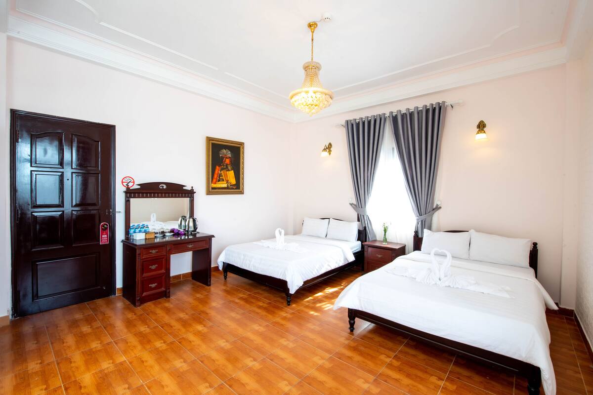 Quarto de hotel com duas camas, cômoda de madeira, escrivaninha com espelho, lustre e um quadro na parede.