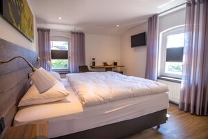 Quarto de hotel moderno com uma cama grande, mesa de cabeceira, escrivaninha e televisão.