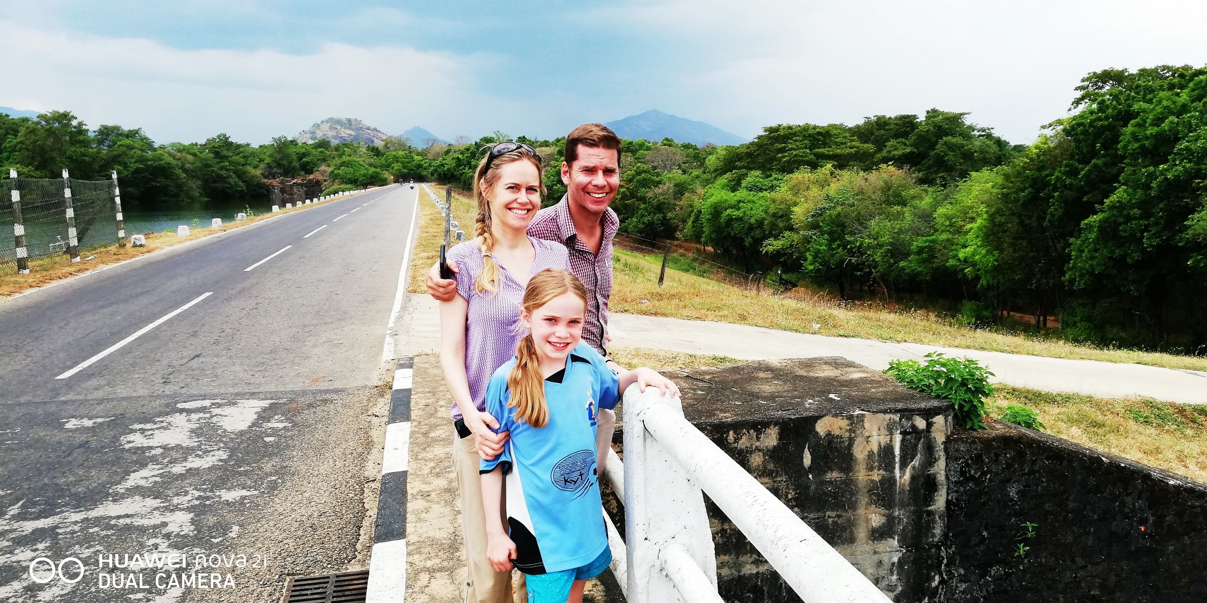 Uma família em uma ponte, com uma estrada e vegetação ao fundo.