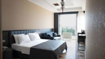 Quarto de hotel moderno com uma cama grande, área de estar com sofá e poltronas, uma mesinha e varanda com vista.
