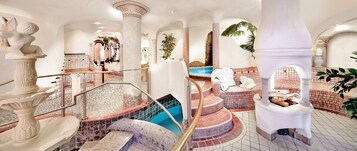 Área de piscina interior luxuosa, com uma banheira de hidromassagem central, vários níveis de escadas e elementos arquitetônicos clássicos como colunas e arcos.