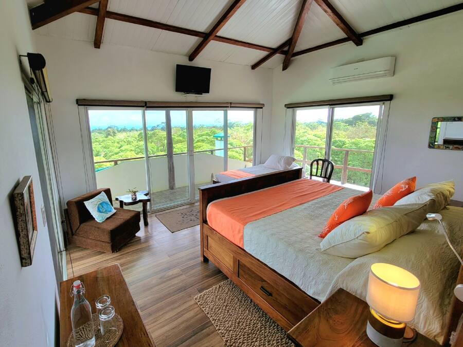 Quarto com cama grande, mesa de cabeceira com abajur, cadeira e vista para o exterior.