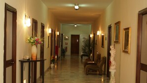 Um corredor com piso de mármore, portas de madeira, luminárias na parede, um vaso com flores e quadros emoldurados.