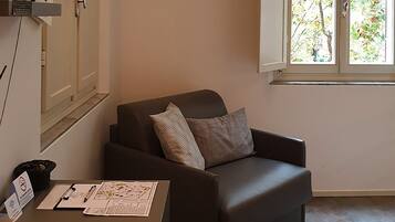 Quarto pequeno e moderno com sofá cinzento, uma cadeira de madeira, escrivaninha com papéis e janela com vista para árvores.
