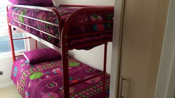 Uma cama beliche com colcha estampada em roxo e verde, uma janela com vista para a rua e uma porta de madeira.