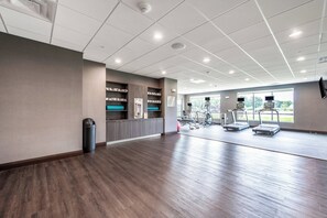 Uma academia espaçosa com piso de madeira, equipamentos de ginástica e uma grande janela que proporciona iluminação natural.