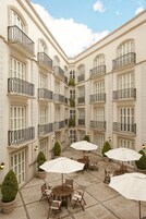 Um pátio com várias mesas e cadeiras, cercado por um edifício com varandas e janelas.