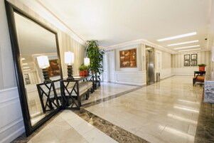 Um hall amplo com piso de mármore, um espelho grande, área de estar com mesa e cadeiras, plantas em vasos e obras de arte emolduradas nas paredes.