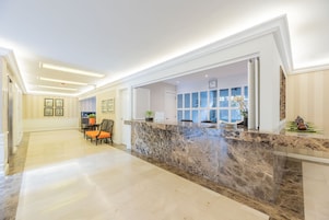 Uma área de recepção com balcão de mármore, espaço de estar com cadeiras e vista do interior pela porta de vidro.