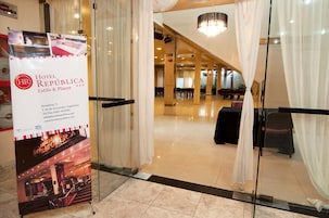 Um saguão de hotel com divisória de vidro, balcão de recepção e escada que leva aos andares superiores.