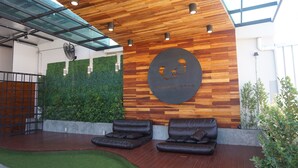Um lobby moderno com painéis de teto de madeira, teto de vidro e uma parede verde com um logotipo circular. Há dois sofás de couro e uma pequena escadaria.