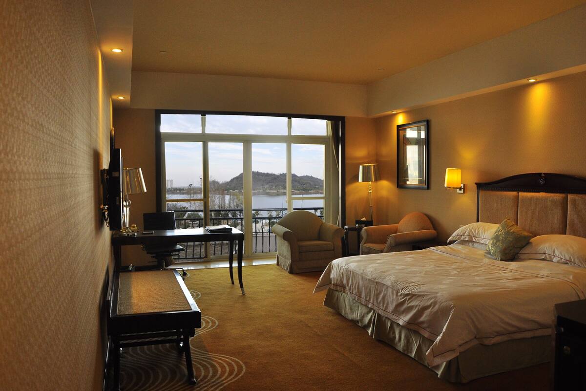 Quarto de hotel com cama, escrivaninha, cadeiras e vista para a água.