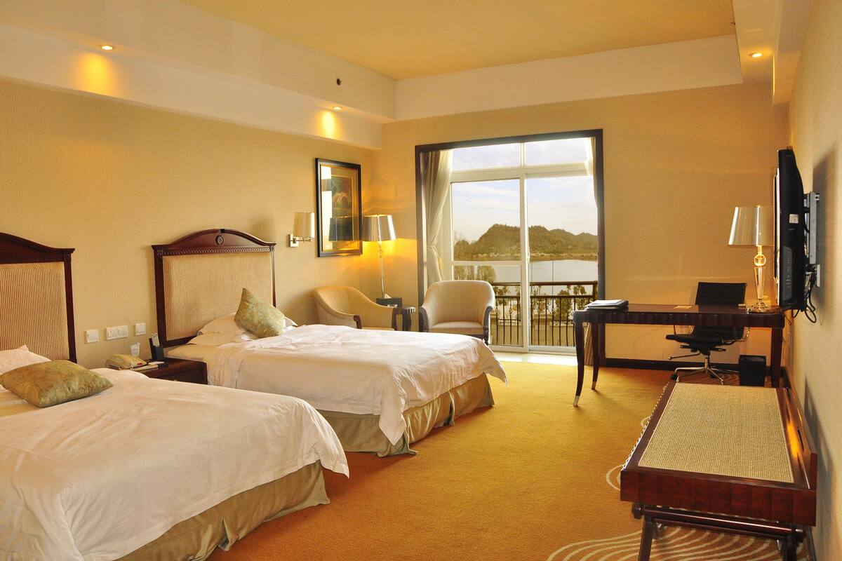 Quarto de hotel com duas camas, uma escrivaninha, uma cadeira, uma televisão e uma grande janela com vista.