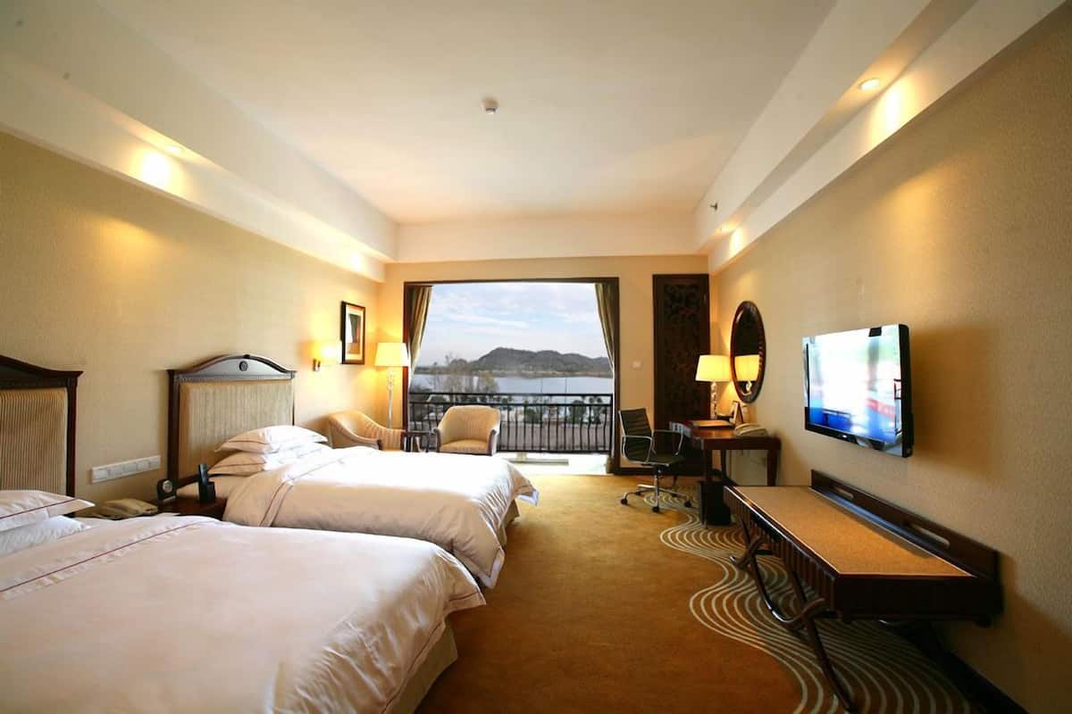 Quarto de hotel com duas camas, uma escrivaninha, uma cadeira, televisão e varanda com vista.