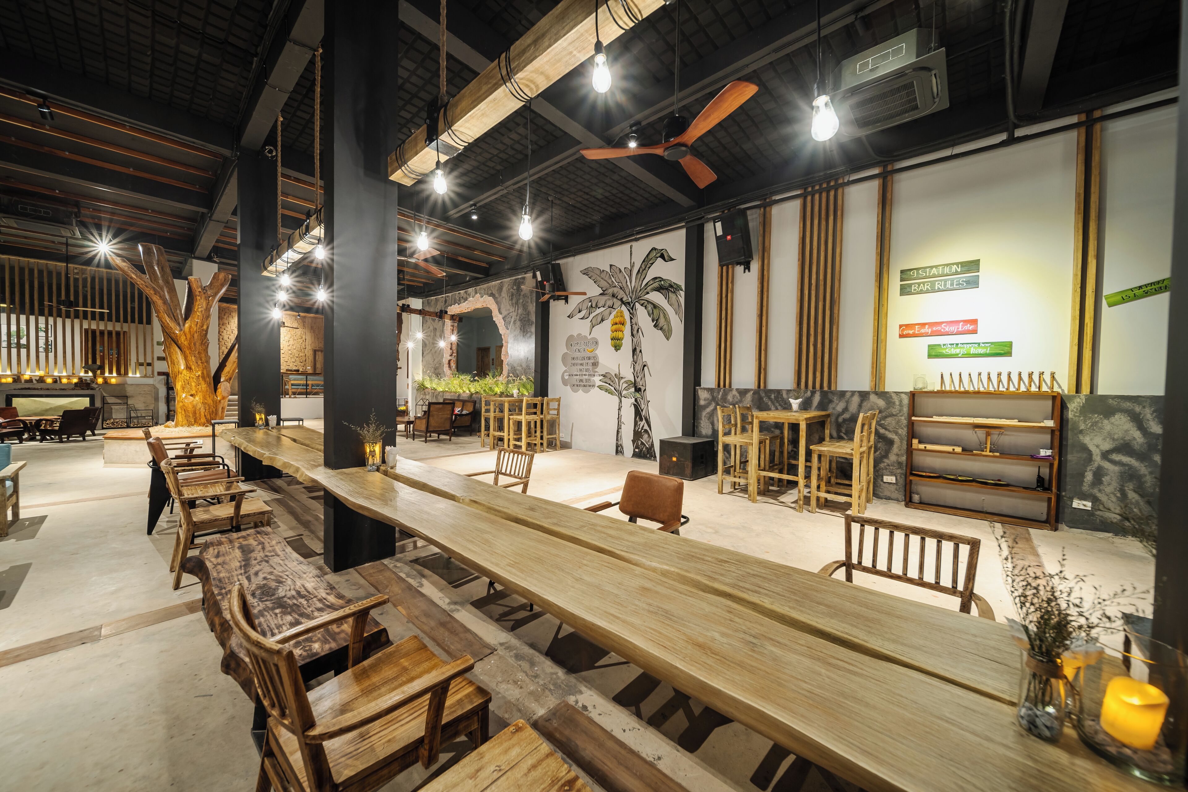 Amplo espaço interior com longas mesas e cadeiras de madeira, estrutura de vigas de madeira e parede com mural e sinalização.