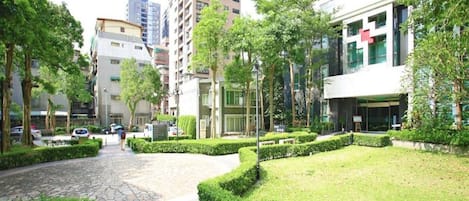 Pátio de hotel bem cuidado, com sebes bem aparadas, um caminho de pedra e edifícios modernos ao fundo.