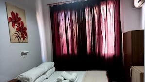 Cama bem arrumada com lençóis e toalhas brancos, mesa de cabeceira de madeira, interruptor de luz fixado na parede e uma grande janela com cortinas vermelho escuro.