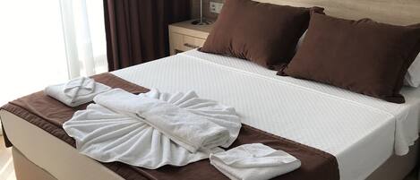 Cama bem arrumada com roupa de cama marrom e branca, cabeceira de madeira, mesa de cabeceira com abajur e janela com cortinas.