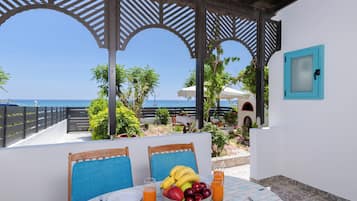 Mesa posta para o café da manhã com frutas e suco de laranja, com vista para uma praia e mar calmo.