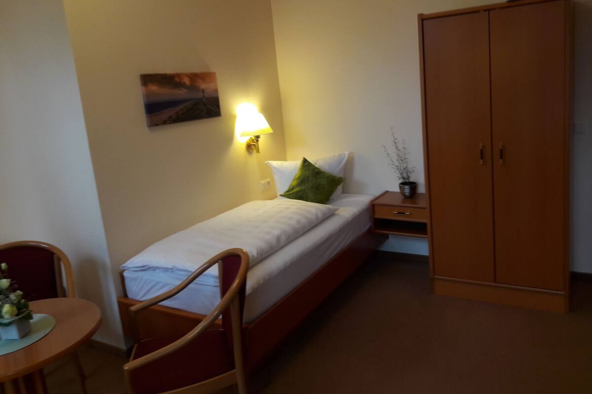 Quarto pequeno e bem arrumado com uma cama de solteiro, mesa de cabeceira com abajur, uma cadeira e um guarda-roupa.