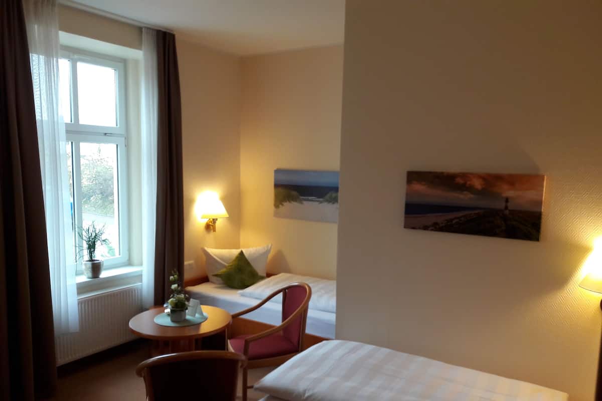 Quarto de hotel com uma cama, uma cadeira, uma mesa pequena com uma planta, uma luminária e um quadro na parede.