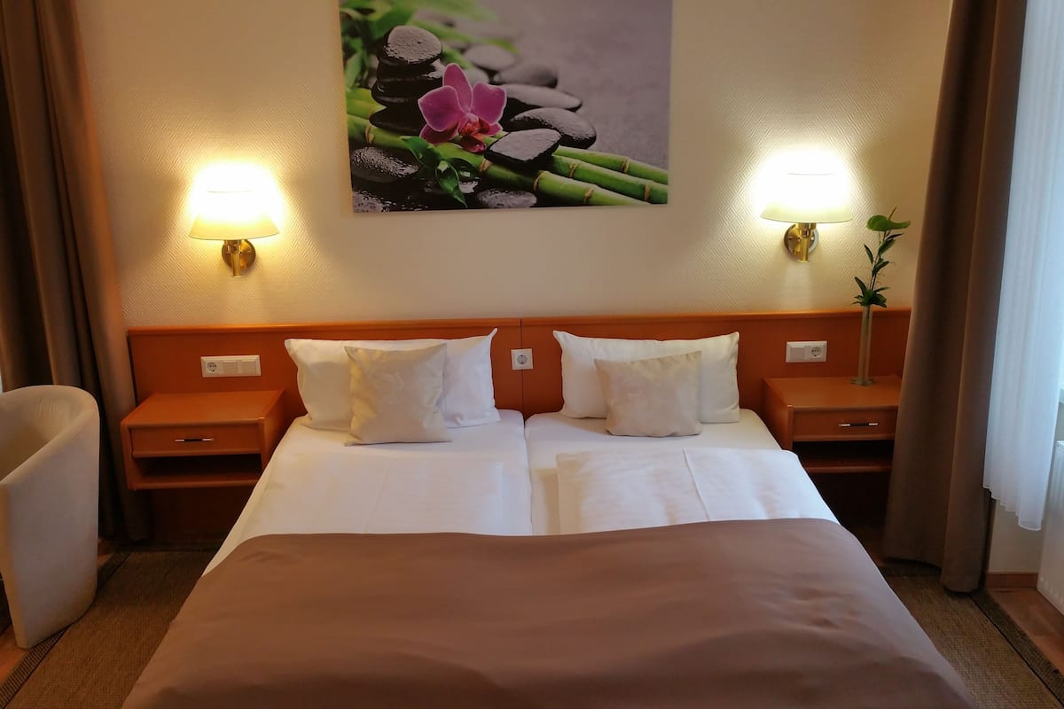 Quarto de hotel com uma cama de casal, duas mesas de cabeceira com luminárias, uma cadeira e um quadro na parede.
