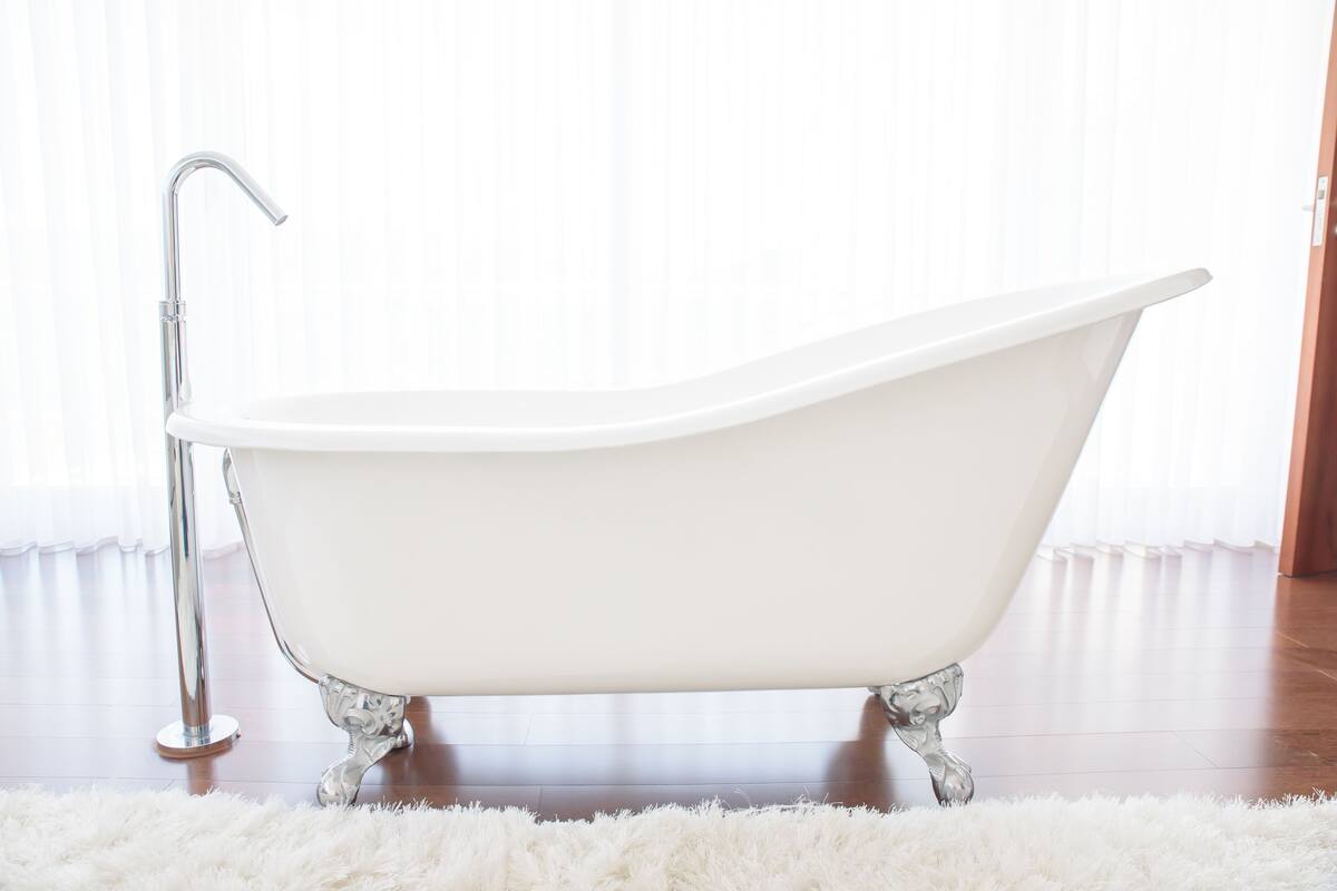 Uma banheira livre, de cor branca, com torneira cromada e pés, sobre um tapete branco e fofo, em um quarto com assoalho de madeira e cortinas leves.