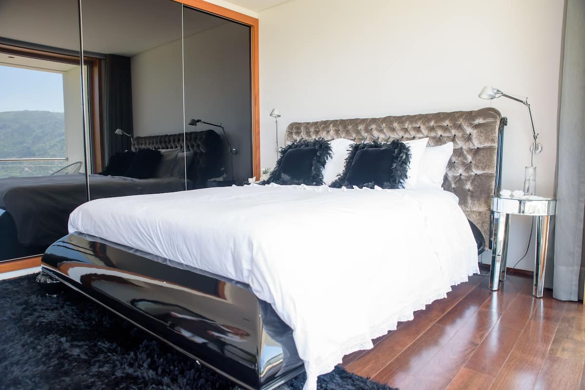 Quarto moderno com cama grande, espelho, mesa de cabeceira e vista para o exterior.