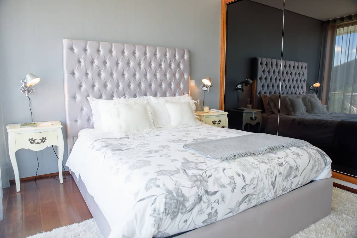 Quarto com cabeceira estofada, colcha florida, duas mesas de cabeceira com abajures e uma janela grande com cortinas.