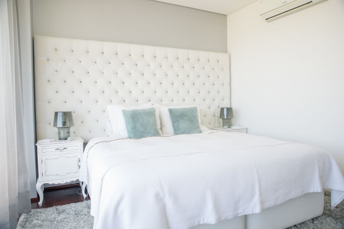 Quarto com cabeceira estofada, edredom branco, duas almofadas, mesa de cabeceira com abajur e cômoda pequena.