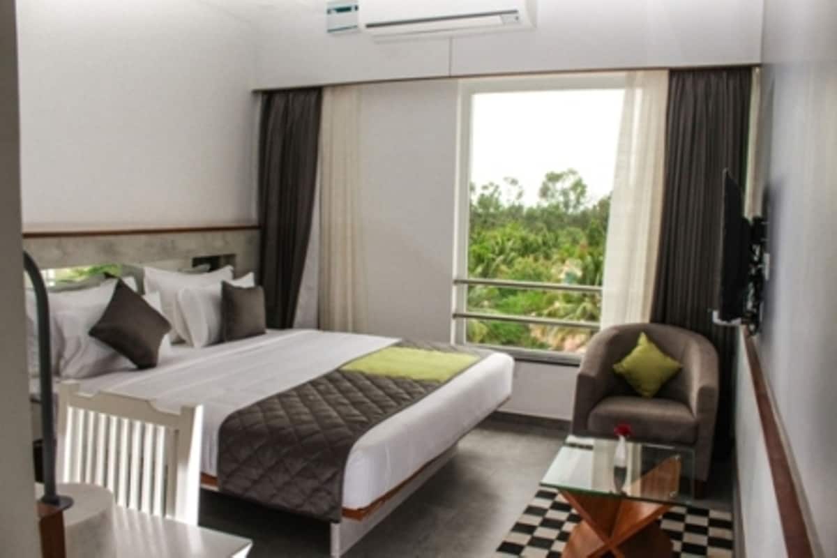 Quarto de hotel com cama, uma cadeira, uma mesinha e janela com vista para o verde.