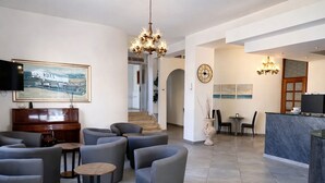 Um hall de hotel com sofás cinzas, um piano de madeira, uma televisão, um candelabro, um relógio e uma mesa de recepção.