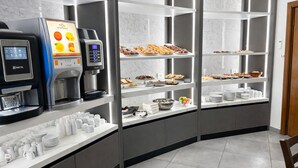 Espaço de buffet de café da manhã com máquina de café, dispensador de bebidas e variedade de pães e frutas.